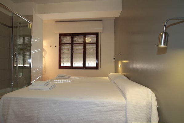 Quarto de hotel com cama, janela com persianas, área de chuveiro e luminária na parede.