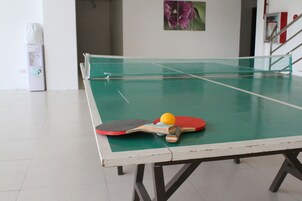 Uma mesa de pingue-pongue com raquetes e uma bola, um dispensador de água e um quadro de flores na parede.