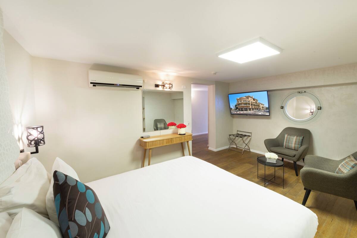 Quarto de hotel com cama, escrivaninha, cadeira, televisão e espelho.