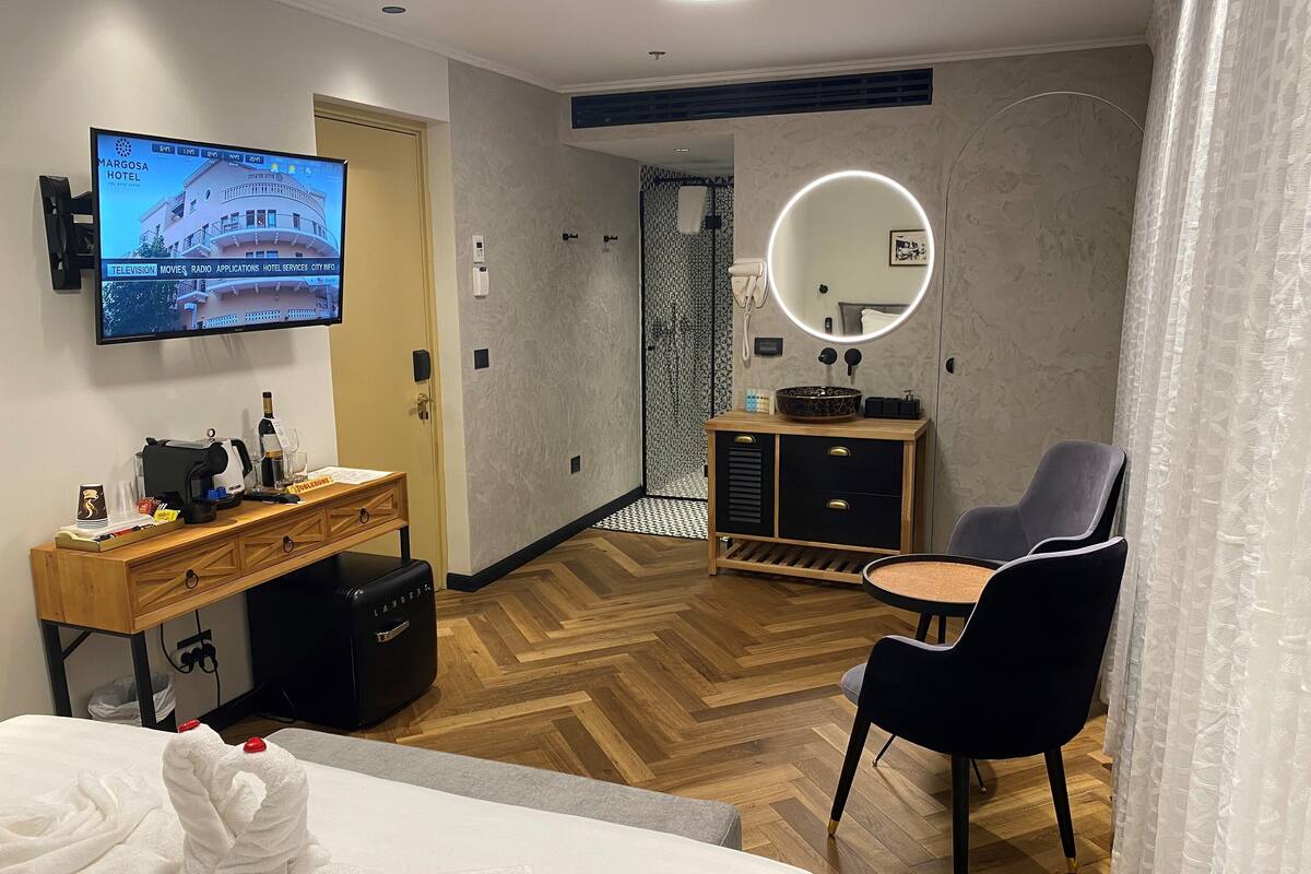 Quarto de hotel com cama, escrivaninha com computador, cadeira, mesa pequena, espelho e televisão exibindo o site do hotel.