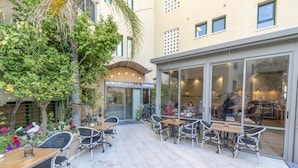 Uma área de café ao ar livre com mesas e cadeiras, um espaço coberto para sentar e a entrada de um edifício com portas de vidro.