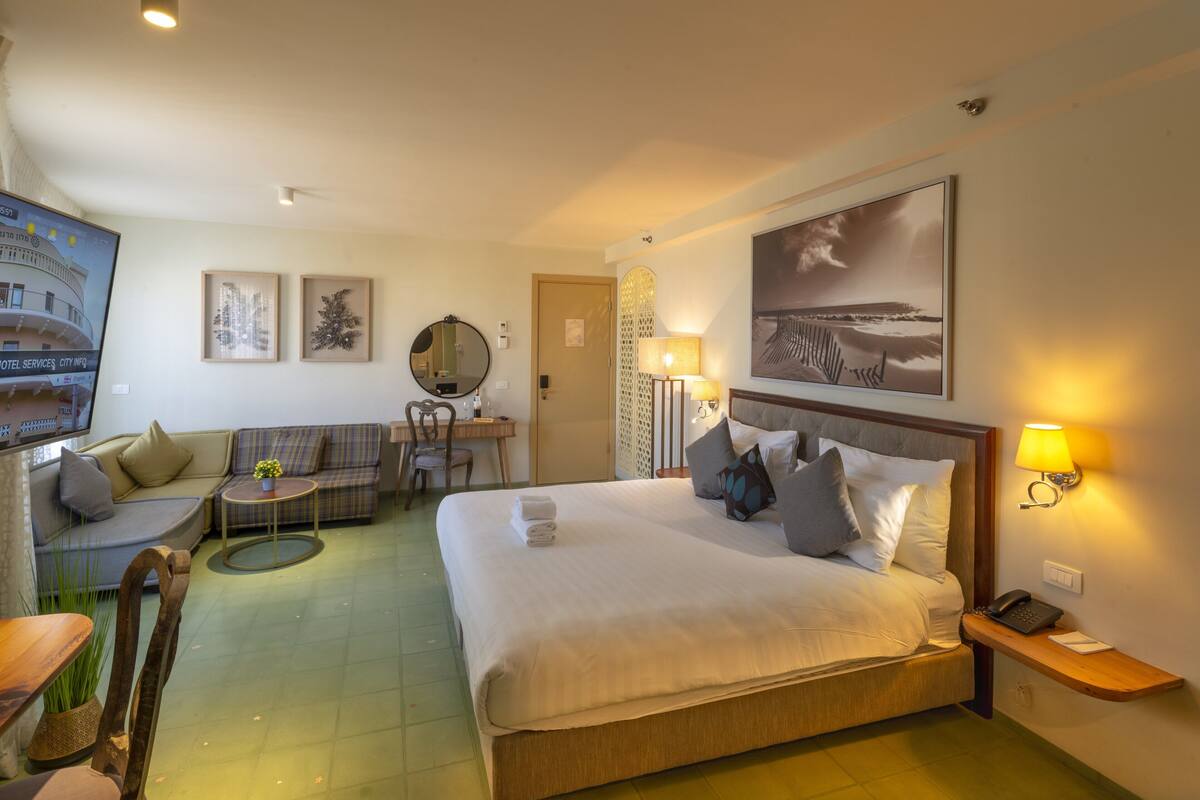 Quarto de hotel com uma cama grande, um sofá, uma mesinha e uma escrivaninha. Há luminárias na parede e um quadro sobre a cama.