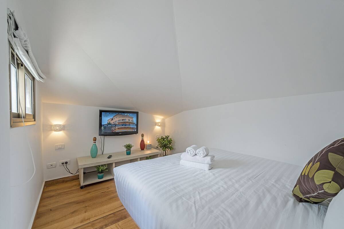 Quarto com cama, televisão na parede, mesa de cabeceira com um vaso e planta, e janela com cortinas.