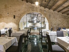 Restaurante com parede de pedra rústica e porta em arco que dá para a cozinha. Mesas com toalhas brancas e cadeiras.