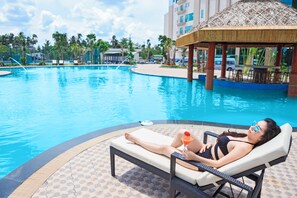 Uma área de estar à beira da piscina com uma pessoa relaxando em um espreguiçadeira, segurando uma bebida.