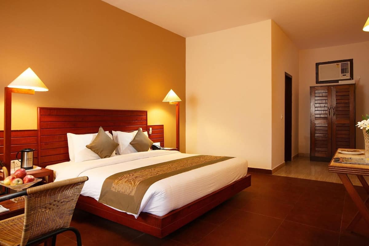 Quarto de hotel com cabeceira de madeira, cama com lençóis brancos, duas luminárias laterais, mesa de cabeceira de madeira com abajur, uma cadeira de vime e uma pequena mesa com uma fruteira.