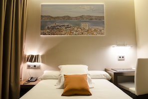 Quarto de hotel com uma cama, mesa de cabeceira com telefone, abajur e pintura de uma cidade costeira.