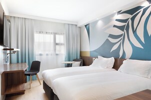 Quarto de hotel com duas camas, uma escrivaninha, uma cadeira, televisão e uma janela grande com cortinas.