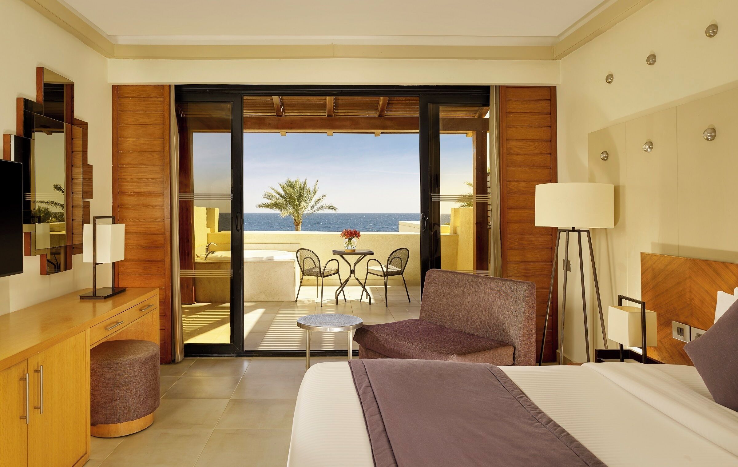 Quarto de hotel com cama, escrivaninha, cadeira, mesinha e varanda com vista para o mar.