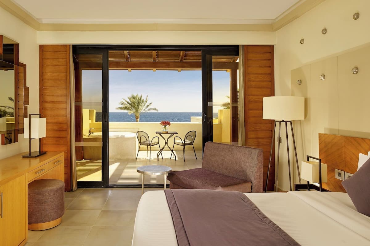 Quarto de hotel com cama, escrivaninha, cadeira, mesinha e varanda com vista para o mar.