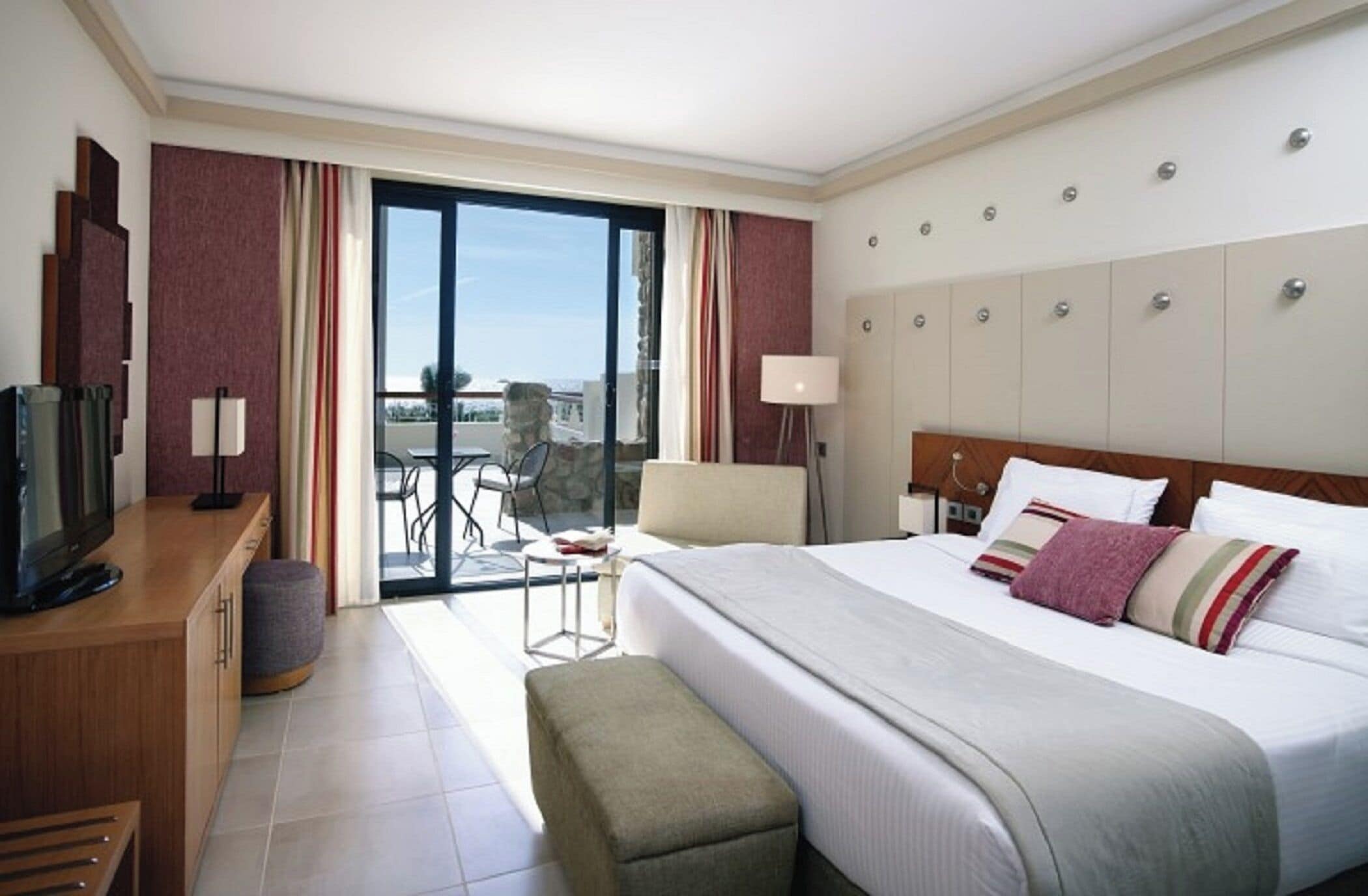 Quarto de hotel com cama, escrivaninha com televisão, poltrona, luminária e vista para o exterior.