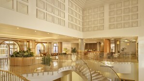 Um amplo hall de hotel com escadaria central, várias áreas de estar e plantas decorativas.