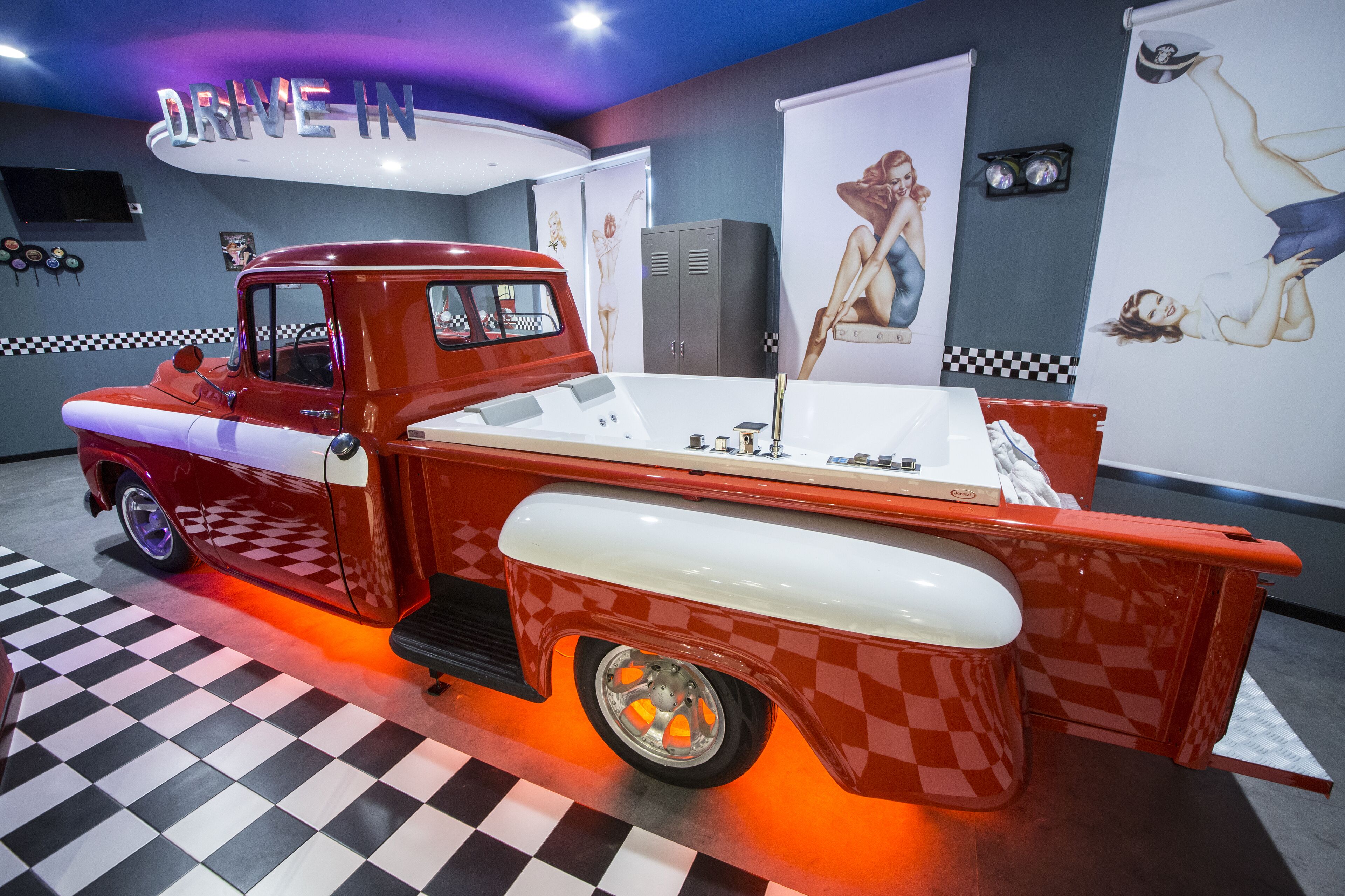Um carro vermelho antigo transformado em banheira em um quarto com piso xadrez, um letreiro 'DRIVE IN' e decoração temática vintage.