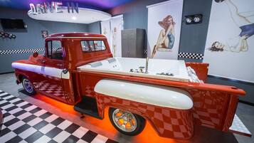 Um carro vermelho antigo transformado em banheira em um quarto com piso xadrez, um letreiro 'DRIVE IN' e decoração temática vintage.