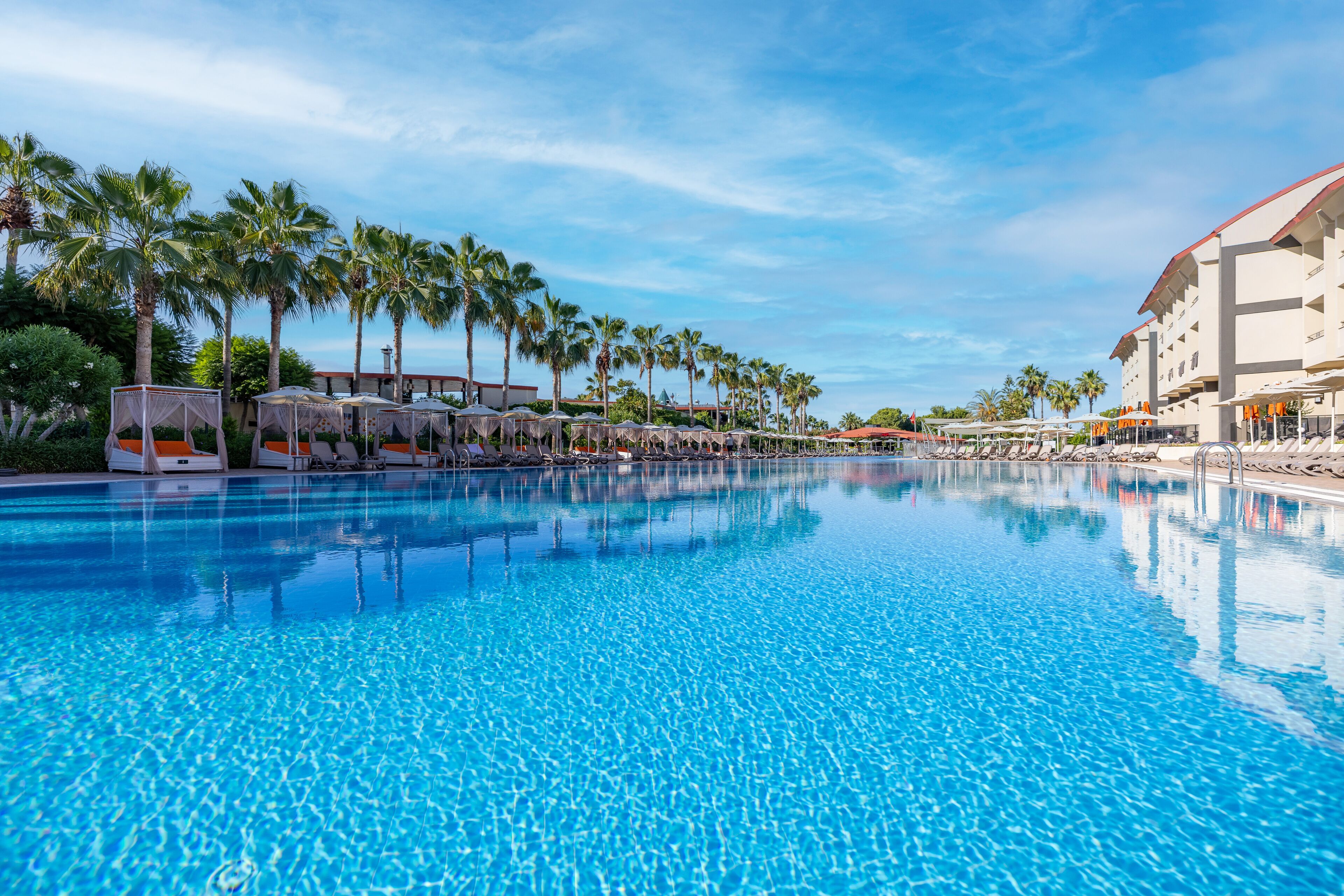 Uma grande piscina azul límpida com espreguiçadeiras e guarda-sóis, cercada por palmeiras e um edifício de hotel.