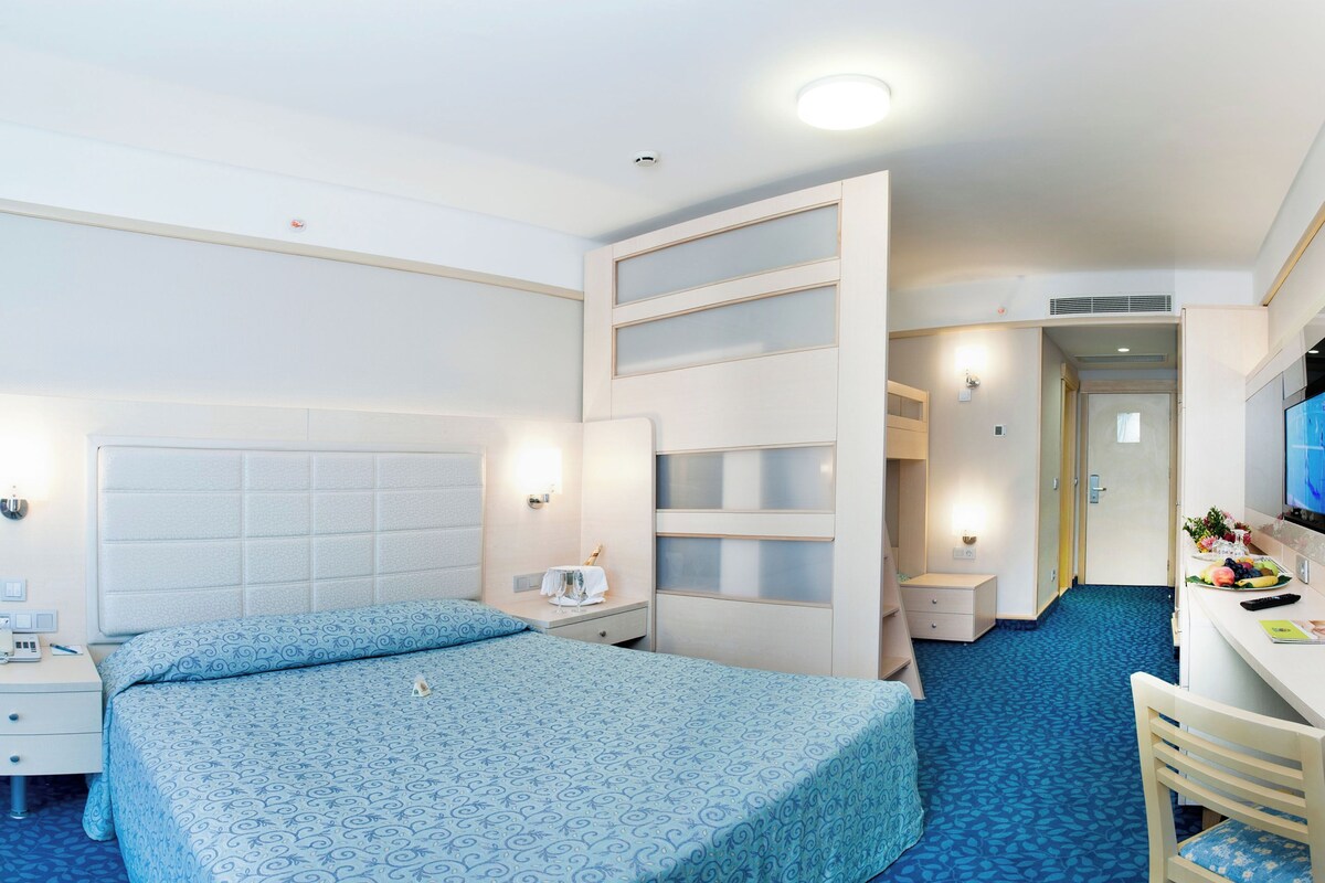 Quarto de hotel com cama, mesa de cabeceira, luminária e escrivaninha com cadeira.
