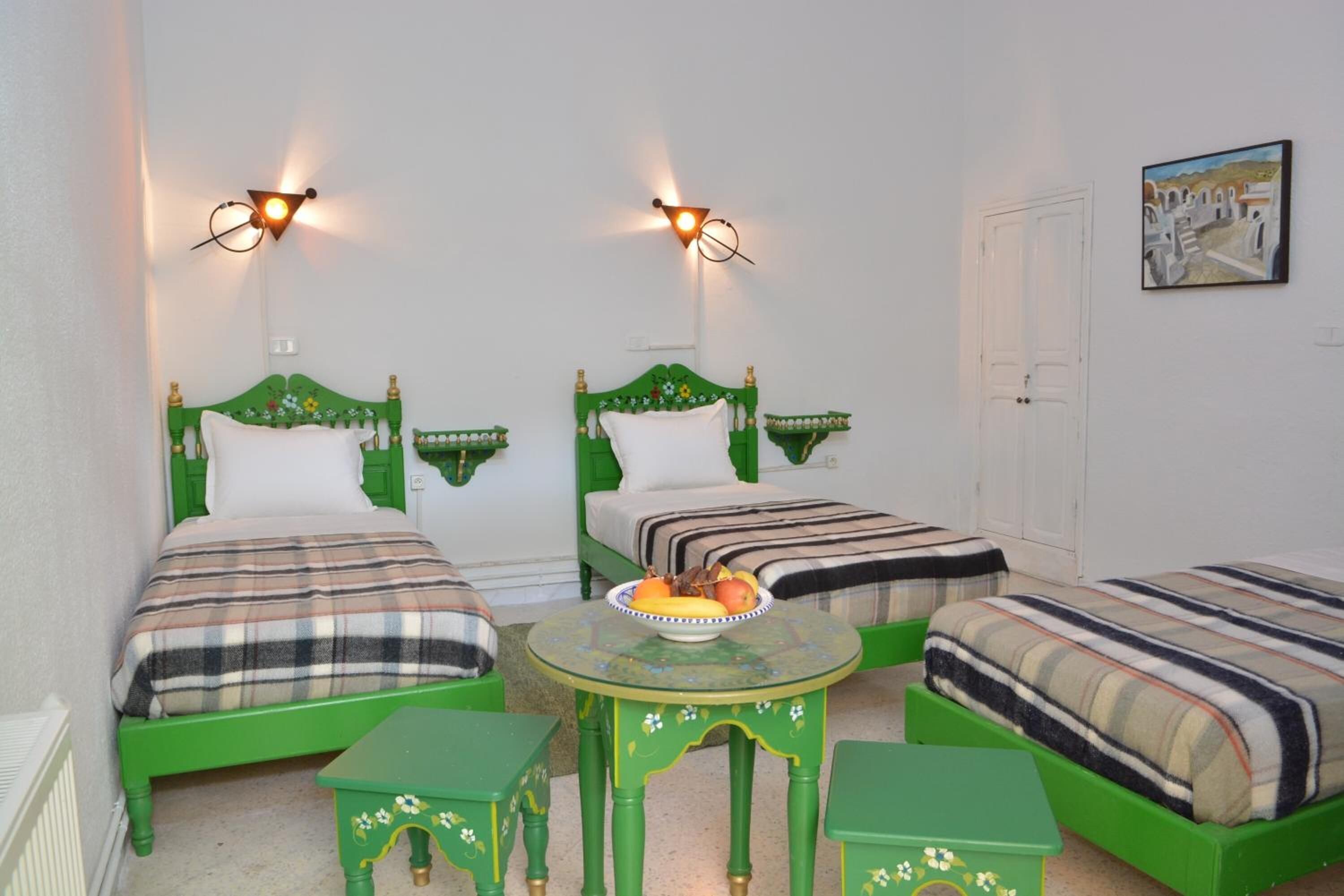Quarto com duas camas verdes, uma mesa redonda com uma tigela de frutas e luminárias fixadas na parede.