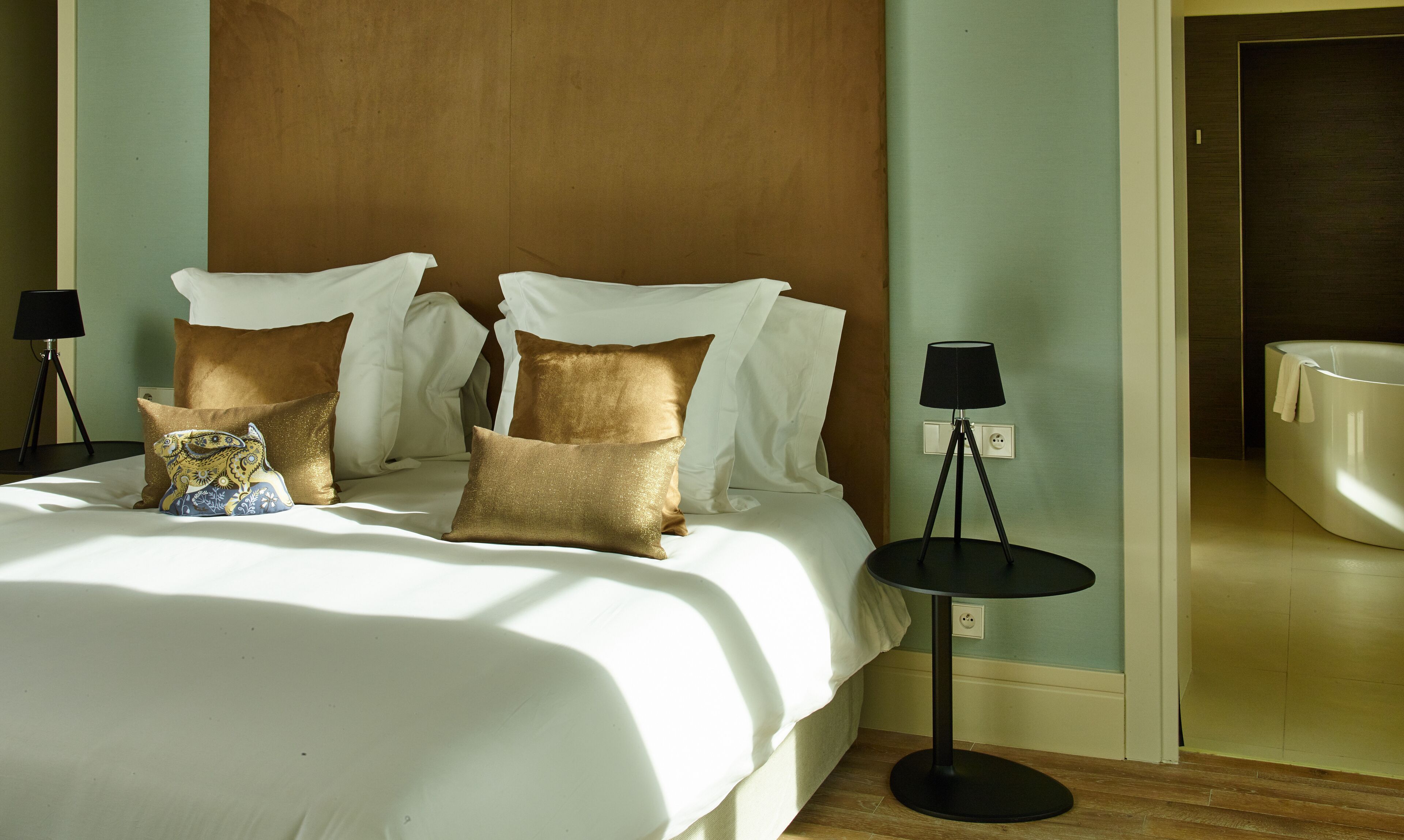 Quarto de hotel com uma cama grande, várias almofadas, mesa de cabeceira com abajur e banheira no banheiro adjacente.