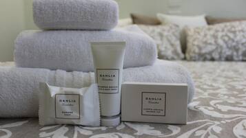Um conjunto de artigos de higiene pessoal organizados, incluindo uma barra de sabão, uma embalagem de champô e um toucabebe, todos da marca Dalia Cosmetics, colocados sobre um edredom estampado.