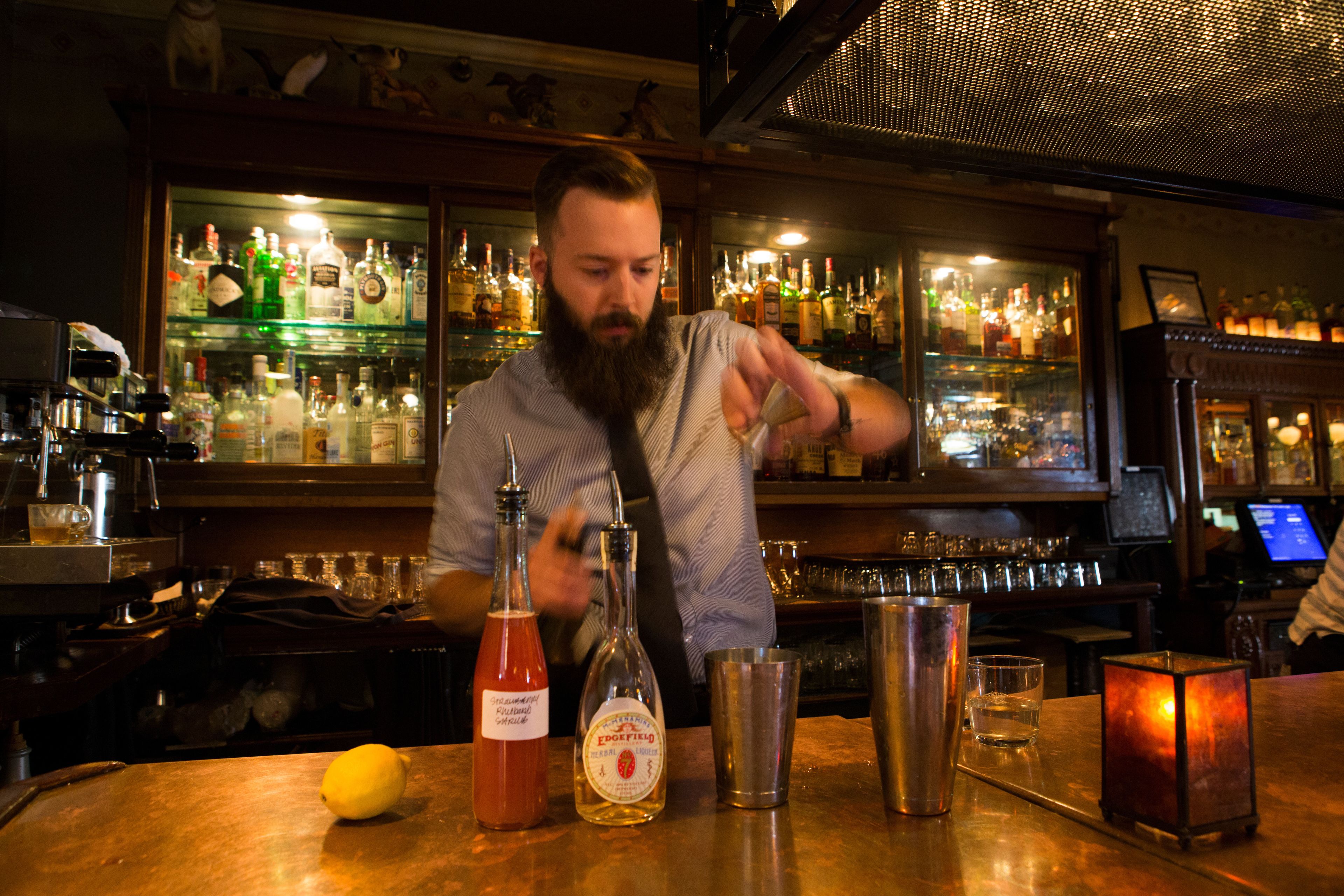 Um bar com um bartender preparando uma bebida. Há várias garrafas e copos nas prateleiras atrás dele.