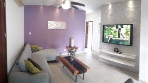 Uma sala de estar com uma parede de destaque roxa, um ventilador de teto, uma TV de tela plana presa a uma parede de pedra, um sofá e uma mesa de centro.