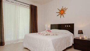 Cama bem arrumada com roupa de cama branca e almofada decorativa, mesa de cabeceira com abajur e um adorno de sol na parede.