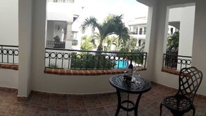 Uma varanda com mesa e cadeira, vista para uma piscina e palmeiras, e um edifício ao fundo.