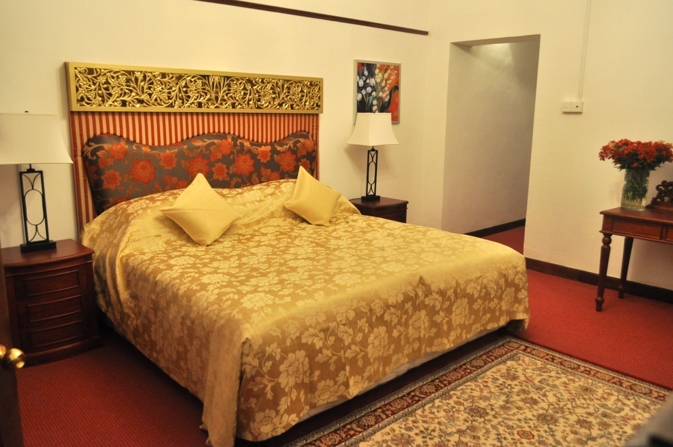 Quarto de hotel com uma cama grande, cabeceira decorativa, mesa de cabeceira com abajur, pequena mesa com um vaso de flores e piso coberto por carpete.