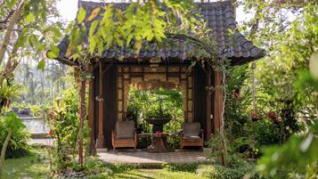Um gazebo de estilo tradicional com telhado de azulejos, rodeado por vegetação exuberante e um pequeno lago.