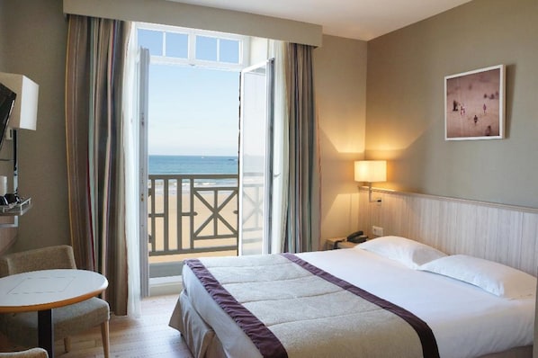 Quarto de hotel com cama, escrivaninha, cadeira, luminária, um quadro e vista para o mar através de uma porta de correr.