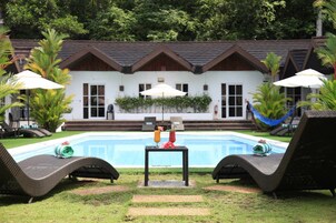 Área de piscina com espreguiçadeiras e guarda-sóis, uma mesa pequena com bebidas, e um edifício com telhado cerâmico ao fundo.