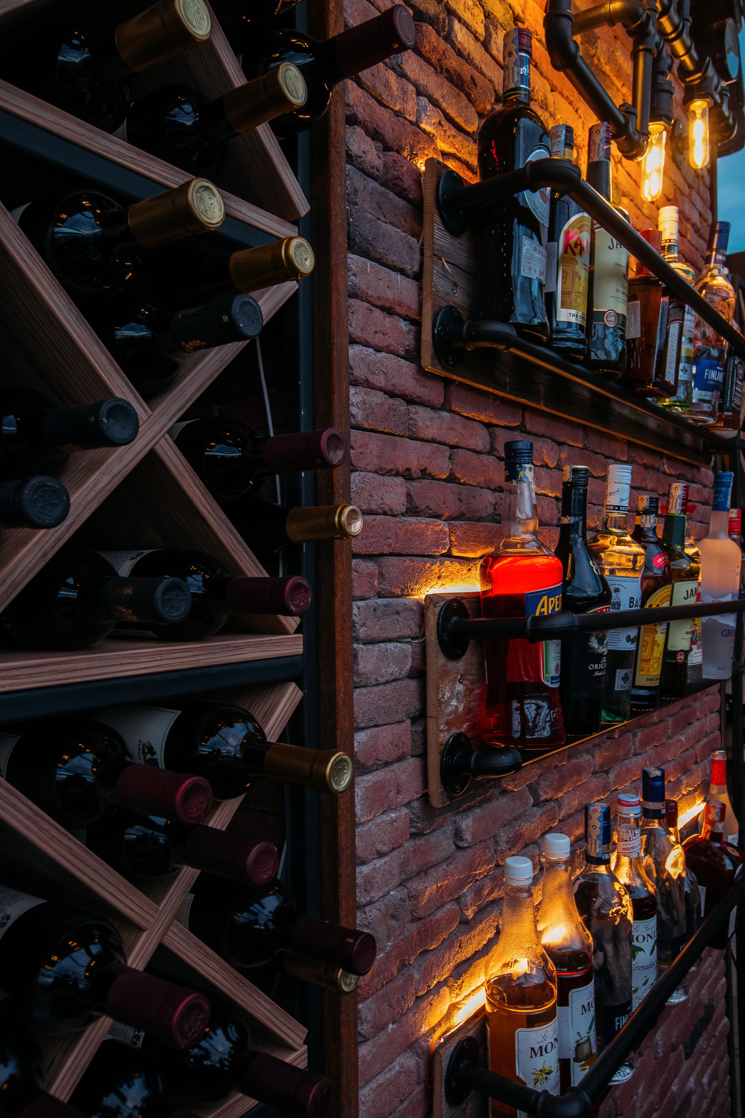 Uma adega com garrafas de vinho e uma prateleira com diversas garrafas de bebidas alcoólicas, tudo em frente a uma parede de tijolos.