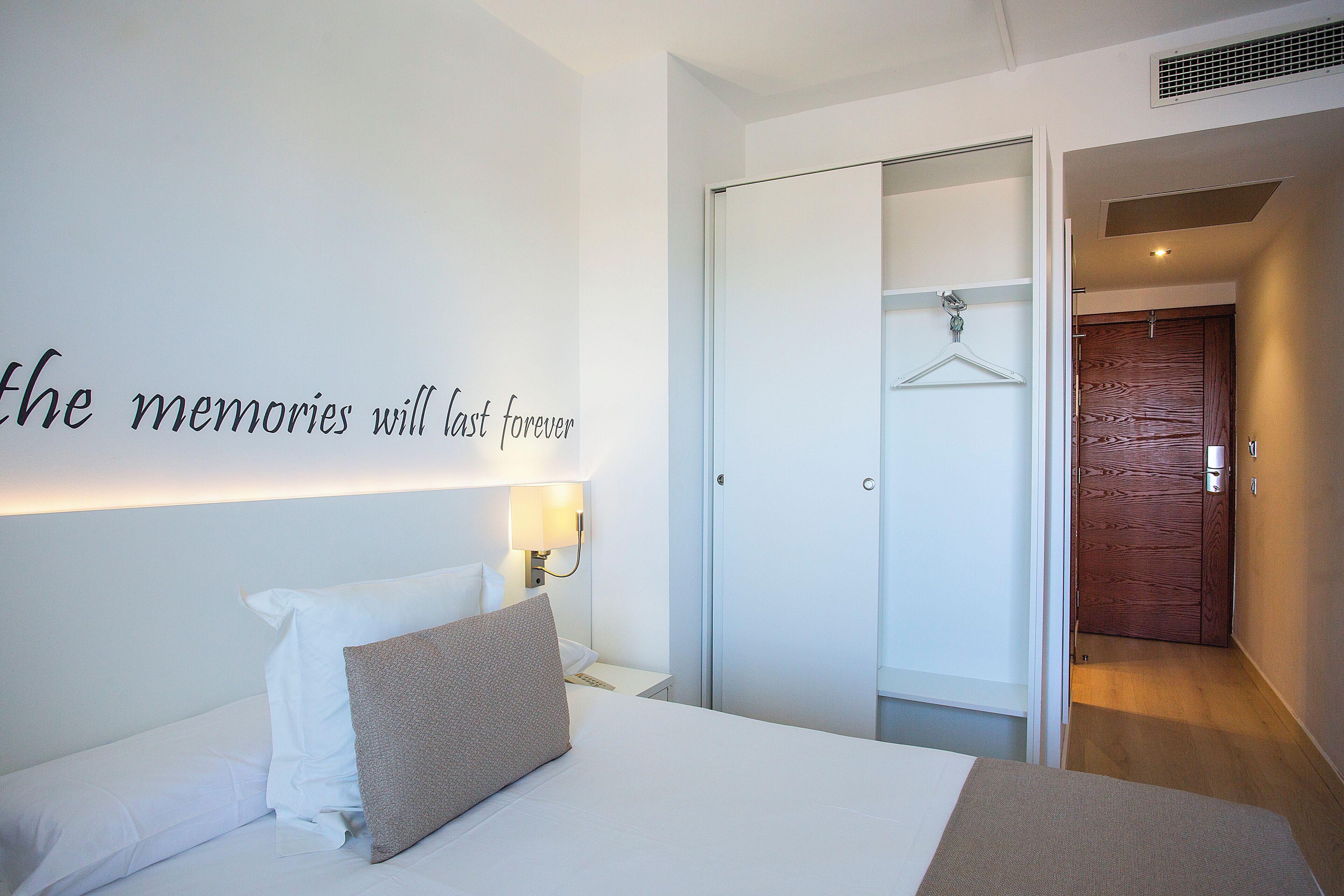 Quarto de hotel com uma cama, mesa de cabeceira, luminária na parede, guarda-roupa e uma porta que dá para outro cômodo.