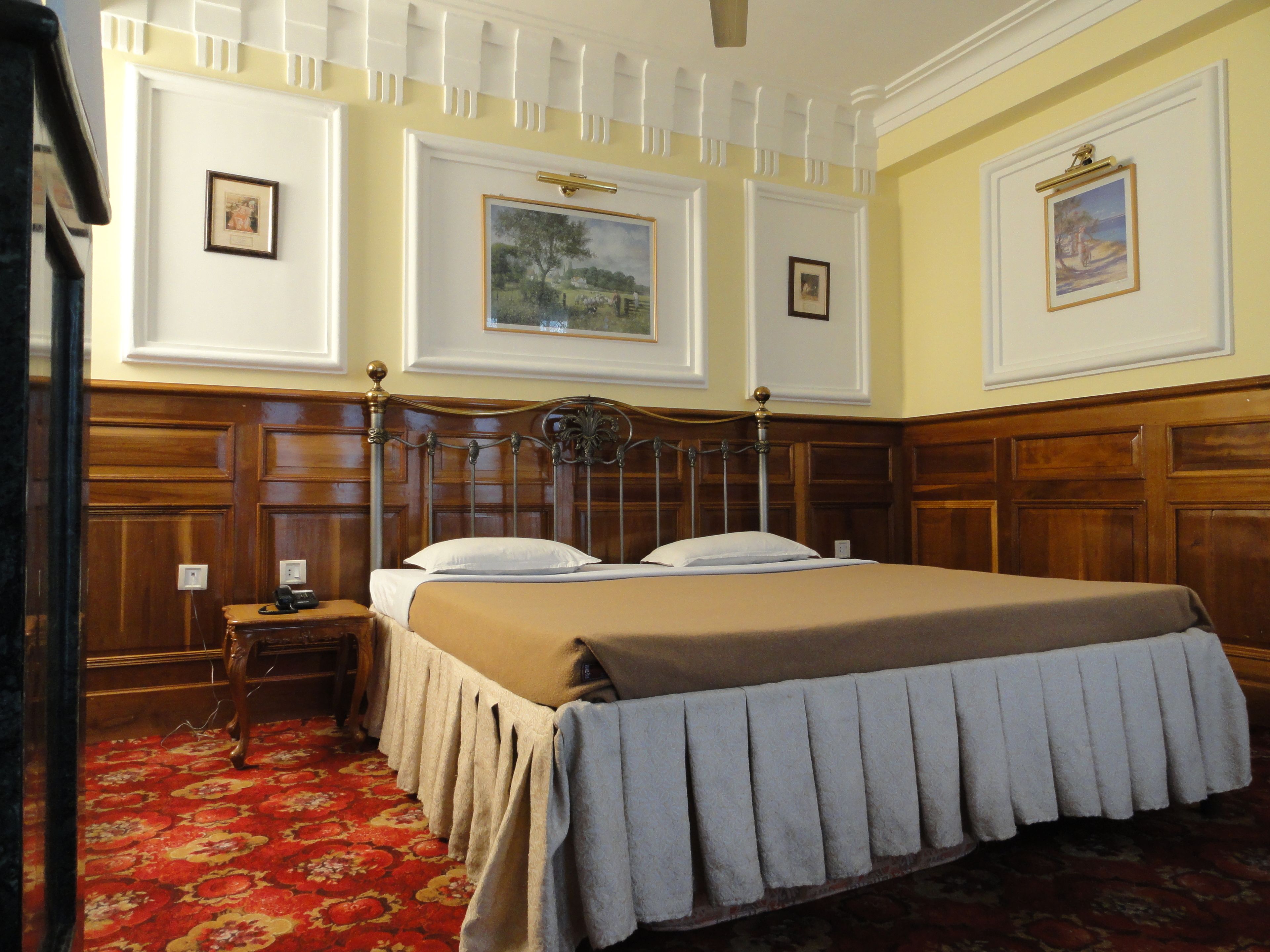 Uma cama bem arrumada com cabeceira de madeira, um tapete floral vermelho e quadros nas paredes.