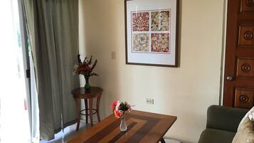 Sala de estar com mesa de centro de madeira, mesinha redonda com um vaso de flores, quadro floral na parede, porta de vidro deslizante e sofá verde.