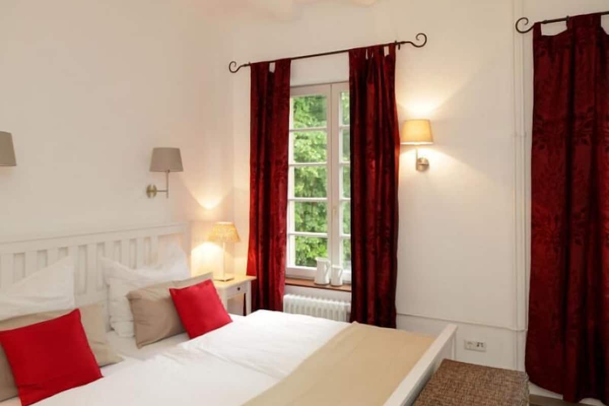 Quarto com cama, duas luminárias de cabeceira, janela com cortinas e radiador.
