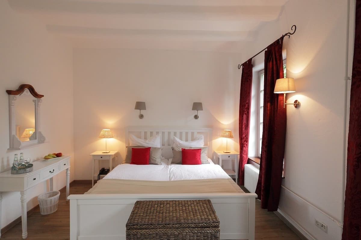 Quarto bem organizado com uma cama grande, duas mesas de cabeceira com abajures, cômoda e um pequeno banquinho de vime.