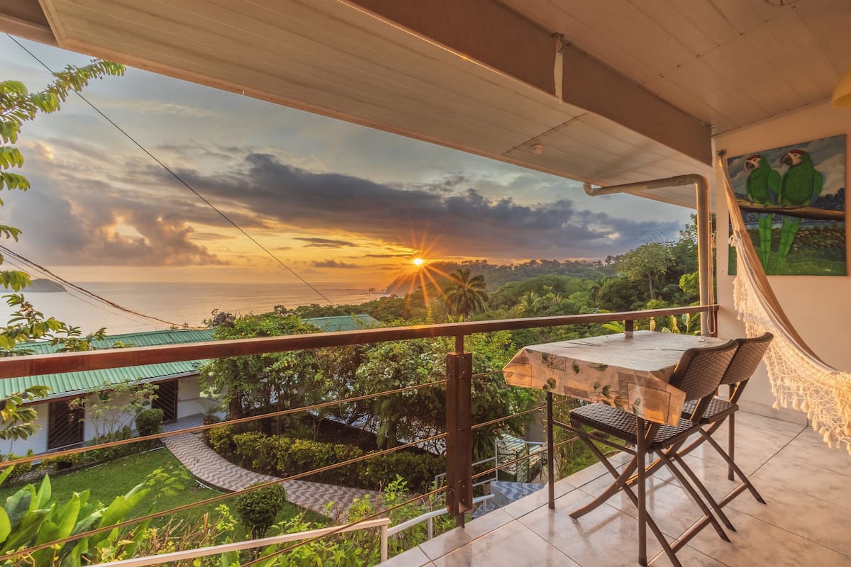 Varanda com mesa e cadeiras, vista para um jardim exuberante e um pôr do sol.