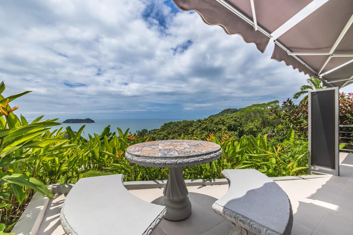 Um pátio com mesa e cadeiras de azulejos, rodeado por vegetação exuberante e vista para o mar.