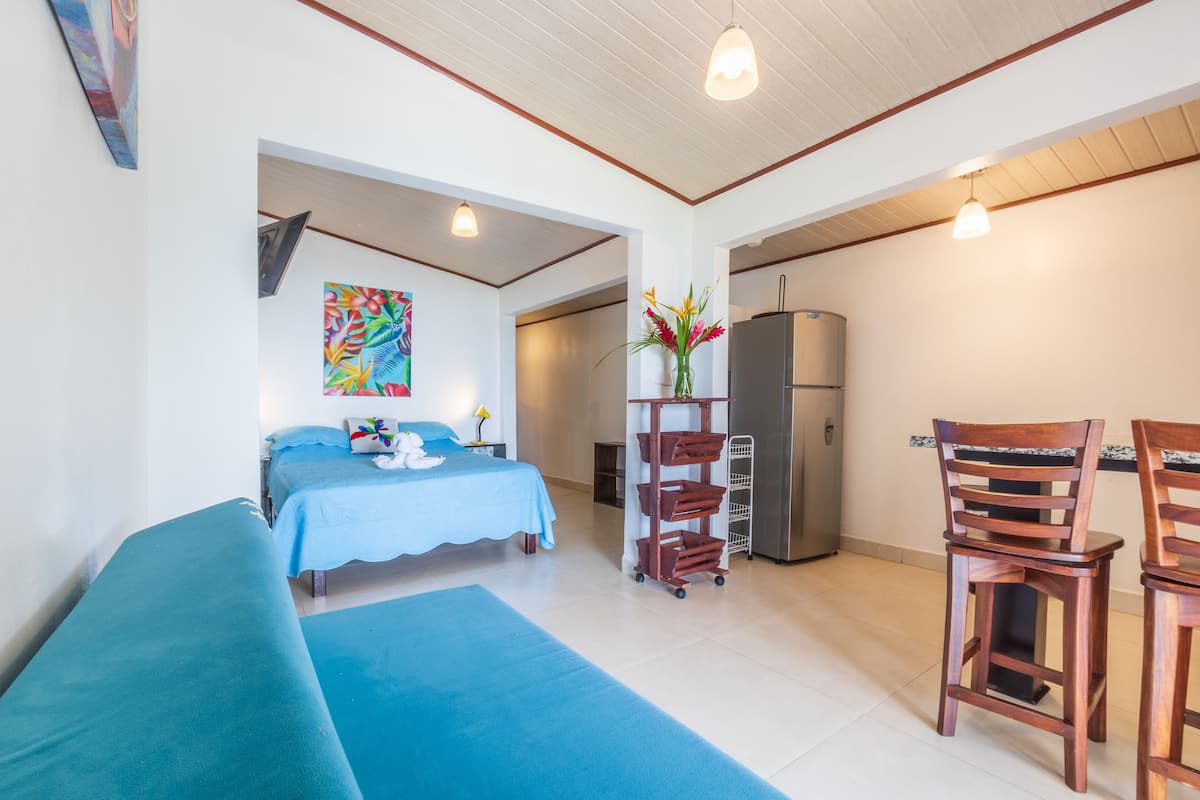 Quarto com cama, sofá azul, cadeira de madeira, mesa pequena e geladeira.