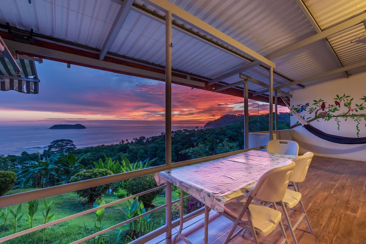 Terraço com mesa e cadeiras, com vista panorâmica para o oceano e vegetação.
