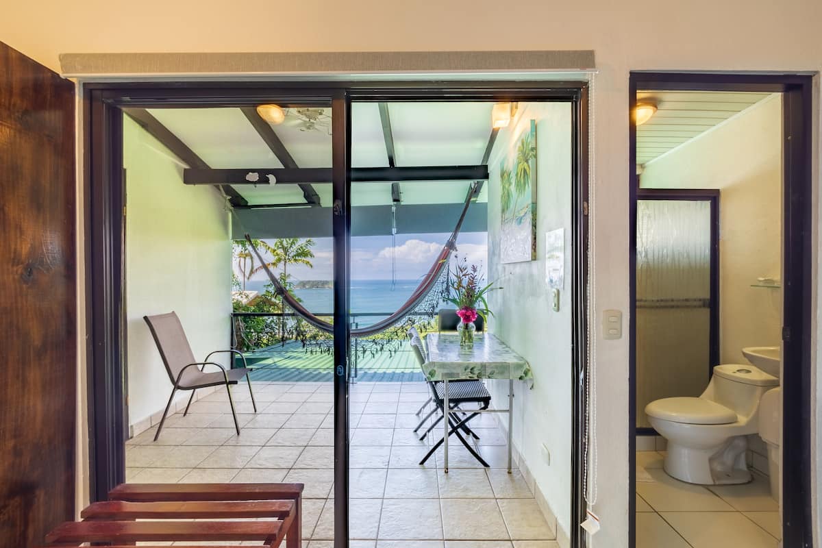 Uma porta deslizante aberta dá para uma varanda com vista para o mar, uma mesa com cadeiras e um banheiro.