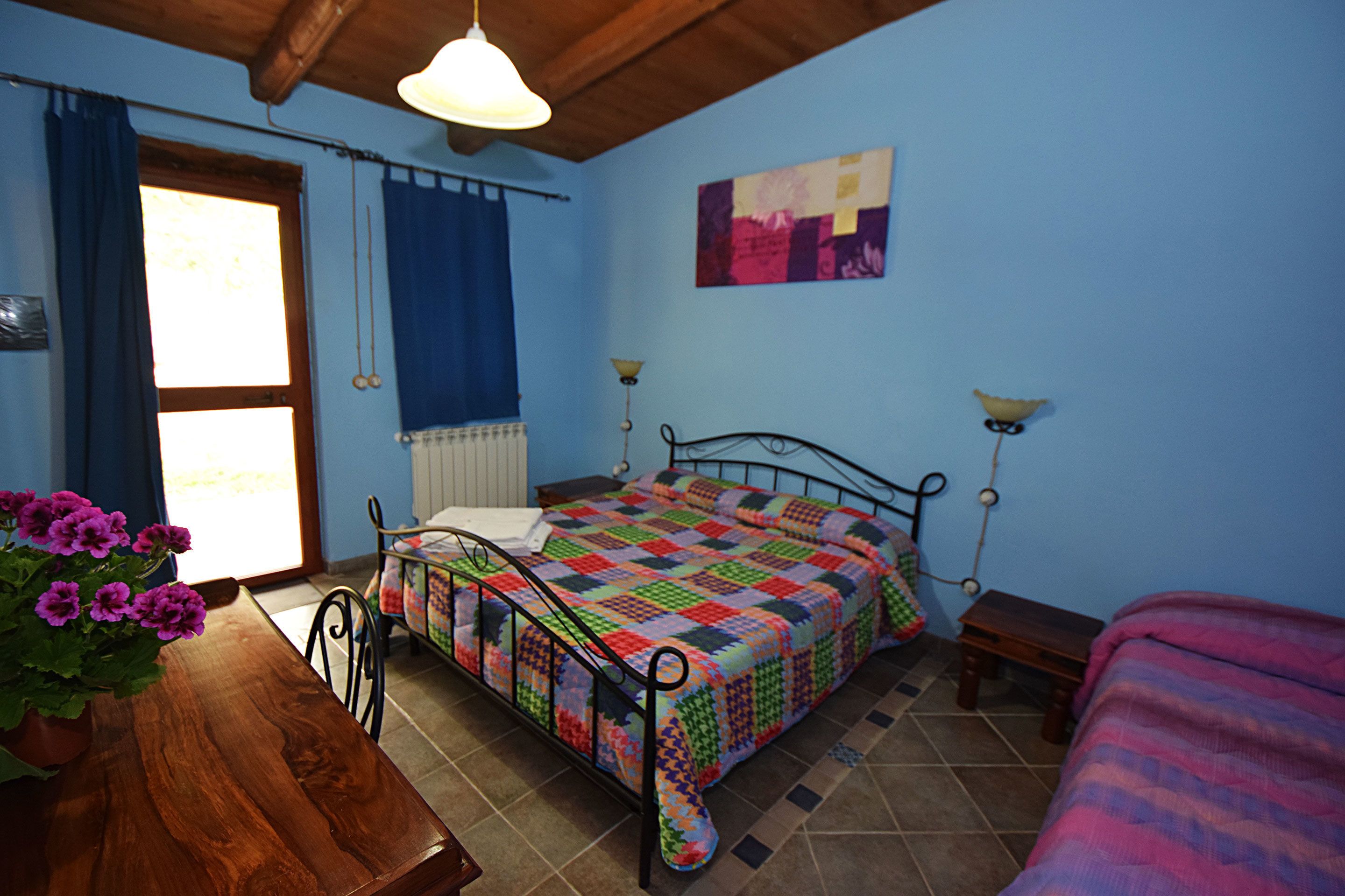 Quarto com cama, mesa de madeira com planta em vaso, janela com cortinas e parede com quadro.