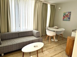 Uma sala de estar com sofá, uma mesa redonda pequena e uma cadeira. Há uma janela com cortinas, um quadro na parede e um armário.