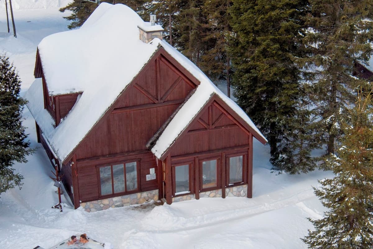 Uma cabana de madeira espaçosa com telhado coberto de neve, uma banheira de hidromassagem e duas motos de neve estacionadas na frente.