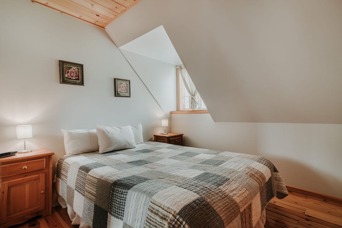 Quarto com teto inclinado, cama com coberta xadrez, duas mesas de cabeceira com luminárias e duas fotos emolduradas na parede.