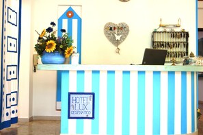 Um balcão de recepção de hotel com padrão azul e branco listrado, uma placa indicando "HOTEL LUX CESENATICO", uma decoração em formato de coração e um suporte de chaves fixado na parede.