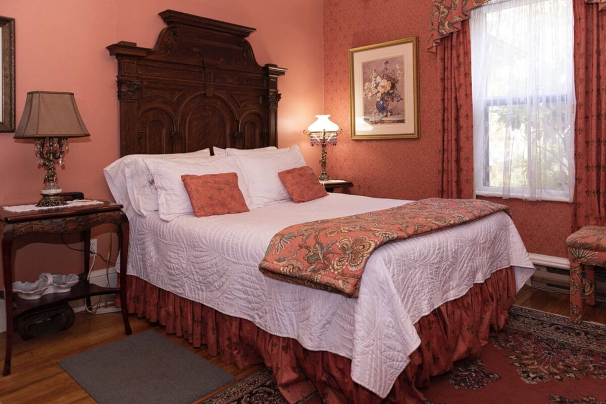 Quarto com uma cama grande, cabeceira de madeira, mesa de cabeceira com abajur, quadro na parede e janela com cortinas.
