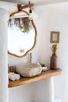 Um banheiro com pia de pedra, penteadeira de madeira, espelho com moldura de madeira, luminária e vaso com plantas secas.