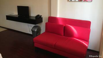 Um sofá vermelho, um suporte de TV preto e uma lixeira num quarto com parede branca e um quadro de um pássaro azul e flores.